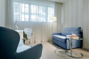 Quarto de hotel moderno com uma poltrona azul, um sofá, uma mesa redonda pequena com uma luminária e vista para a cidade através de grandes janelas.