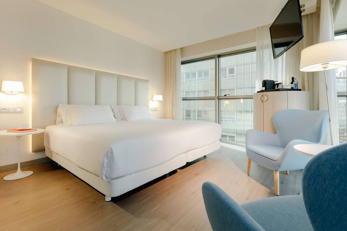 Quarto de hotel moderno com uma cama grande, área de estar com uma poltrona e uma mesinha, televisão fixada na parede e uma janela ampla com vista para a cidade.