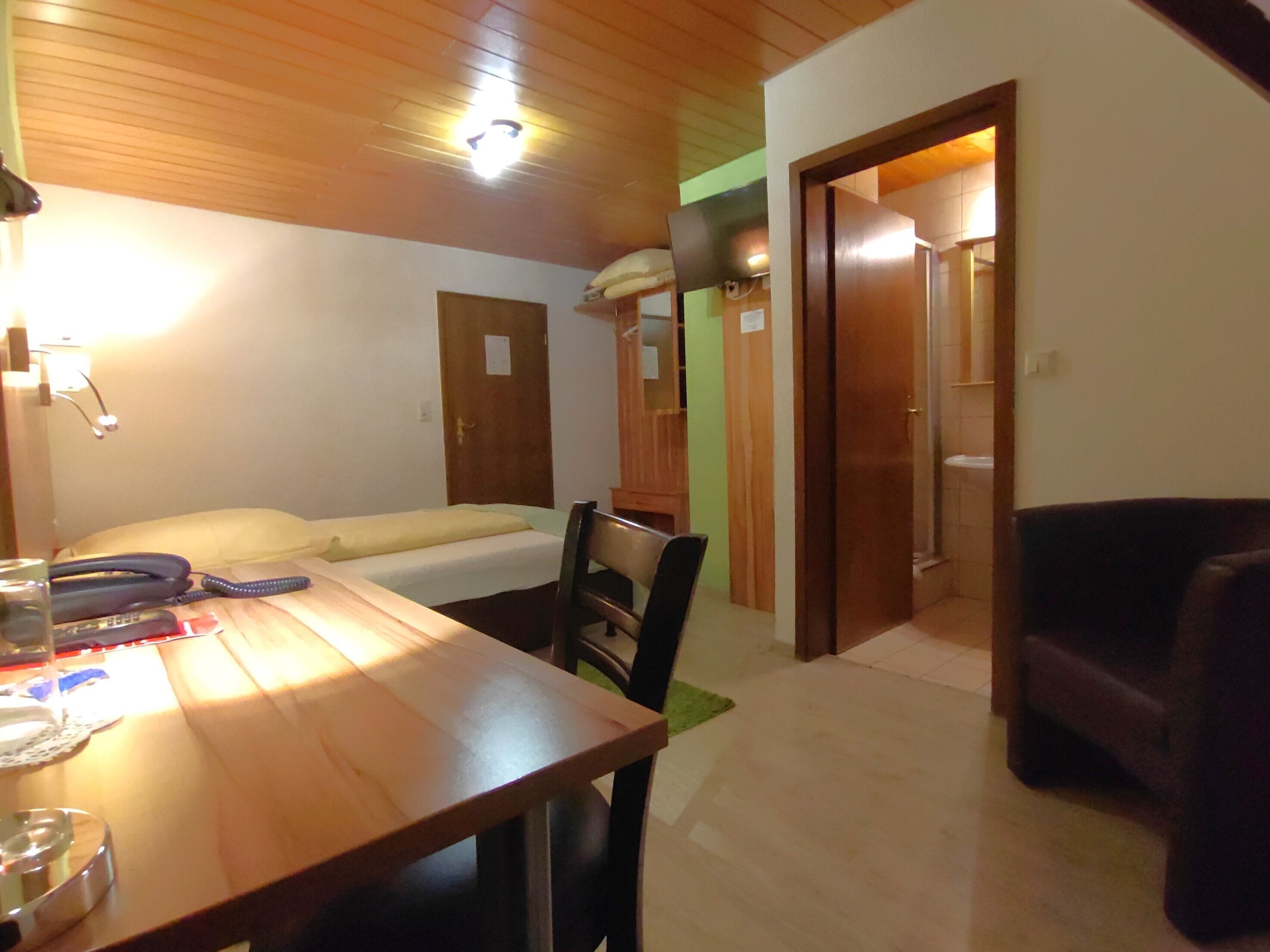 Quarto com teto de madeira, uma cama, uma mesa com cadeiras e uma porta que dá para outro cômodo.