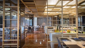 Um restaurante moderno com layout aberto e arejado, com vigas de madeira e divisórias de vidro. As mesas estão bem organizadas com cadeiras, e uma área de buffet é visível ao fundo.