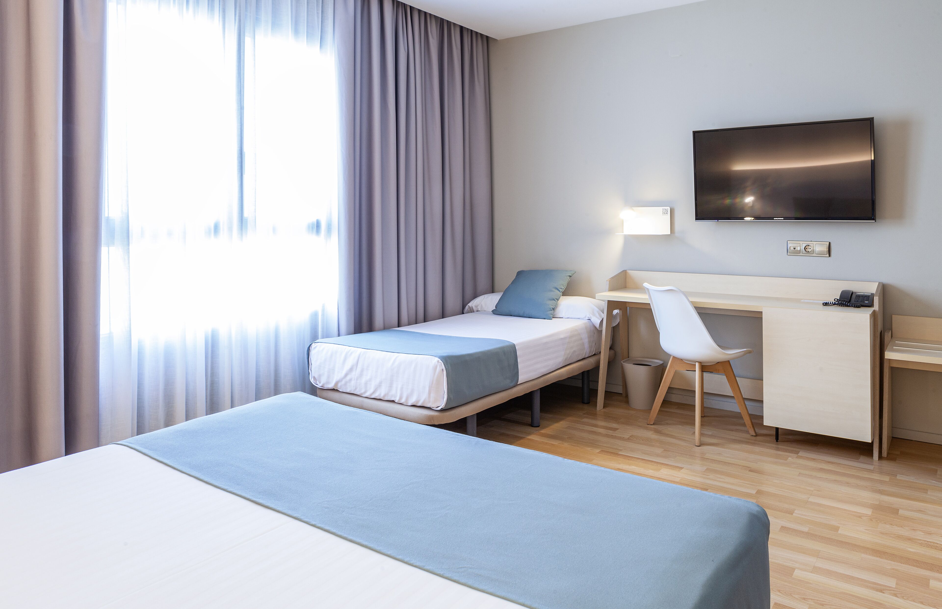 Quarto de hotel com cama, escrivaninha com cadeira, televisão e janela com cortinas.