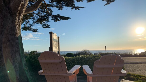 Duas cadeiras Adirondack de frente para uma vista panorâmica do oceano ao pôr do sol, com uma placa de madeira ao fundo.