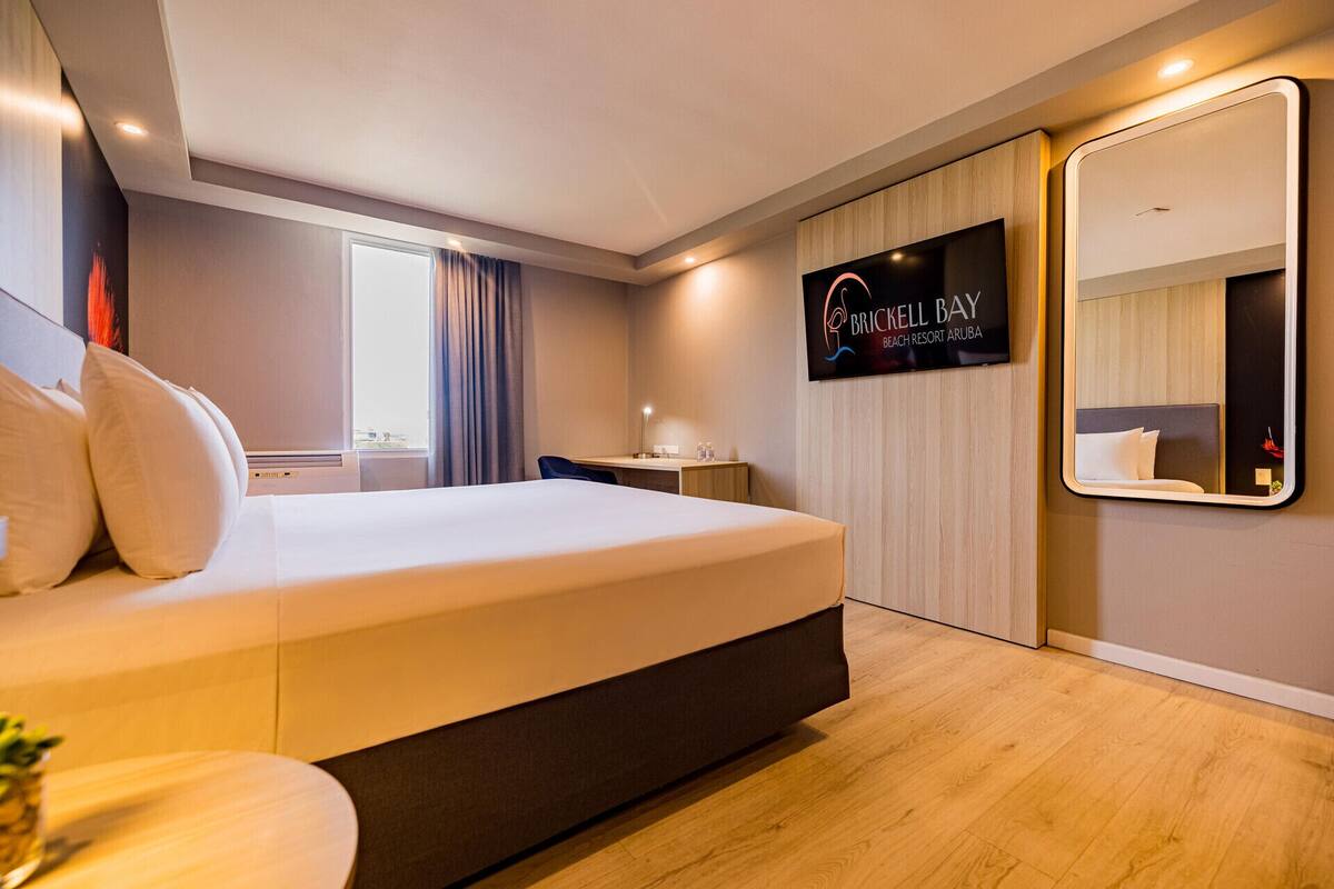 Quarto de hotel com cama, escrivaninha, cadeira, televisão e espelho.