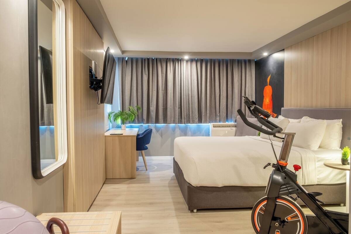Quarto de hotel moderno com cama, bicicleta ergométrica, escrivaninha, cadeira e televisão na parede.