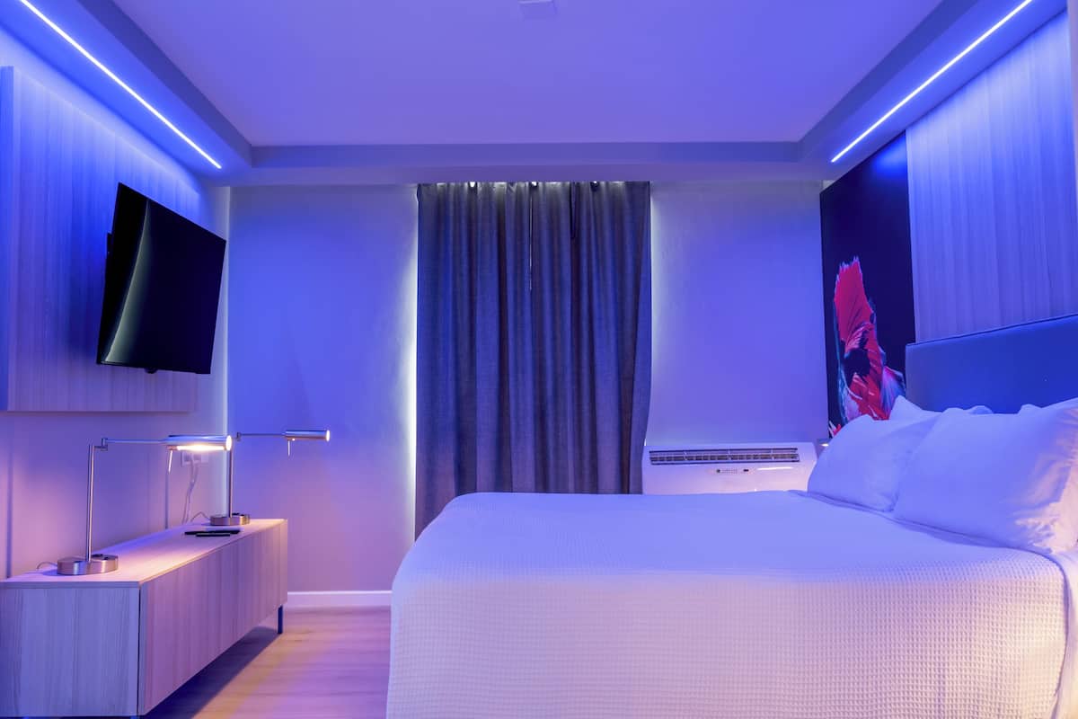 Quarto de hotel moderno com uma cama grande, televisão de tela plana na parede, mesa de cabeceira com abajur e uma janela com cortinas.