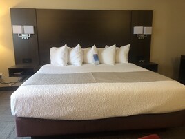 Quarto de hotel com uma cama grande, várias almofadas, mesas de cabeceira com abajures e um criado-mudo com relógio.