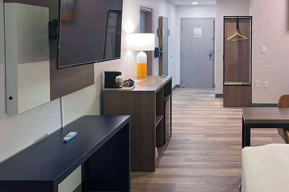 Um corredor de hotel com uma mesa, uma lâmpada, um porta-roupas e uma porta no final.