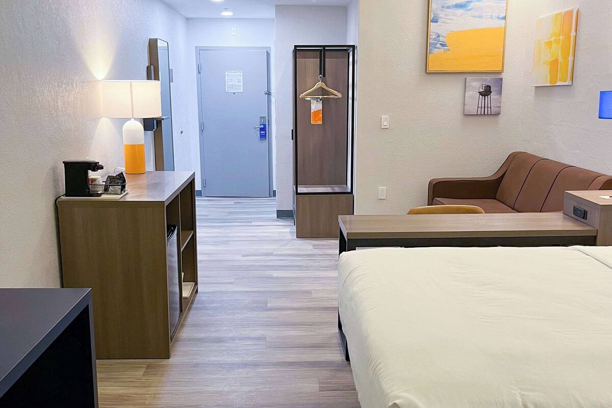 Quarto de hotel com cama, escrivaninha com luminária, cômoda, sofá e cabideiro.