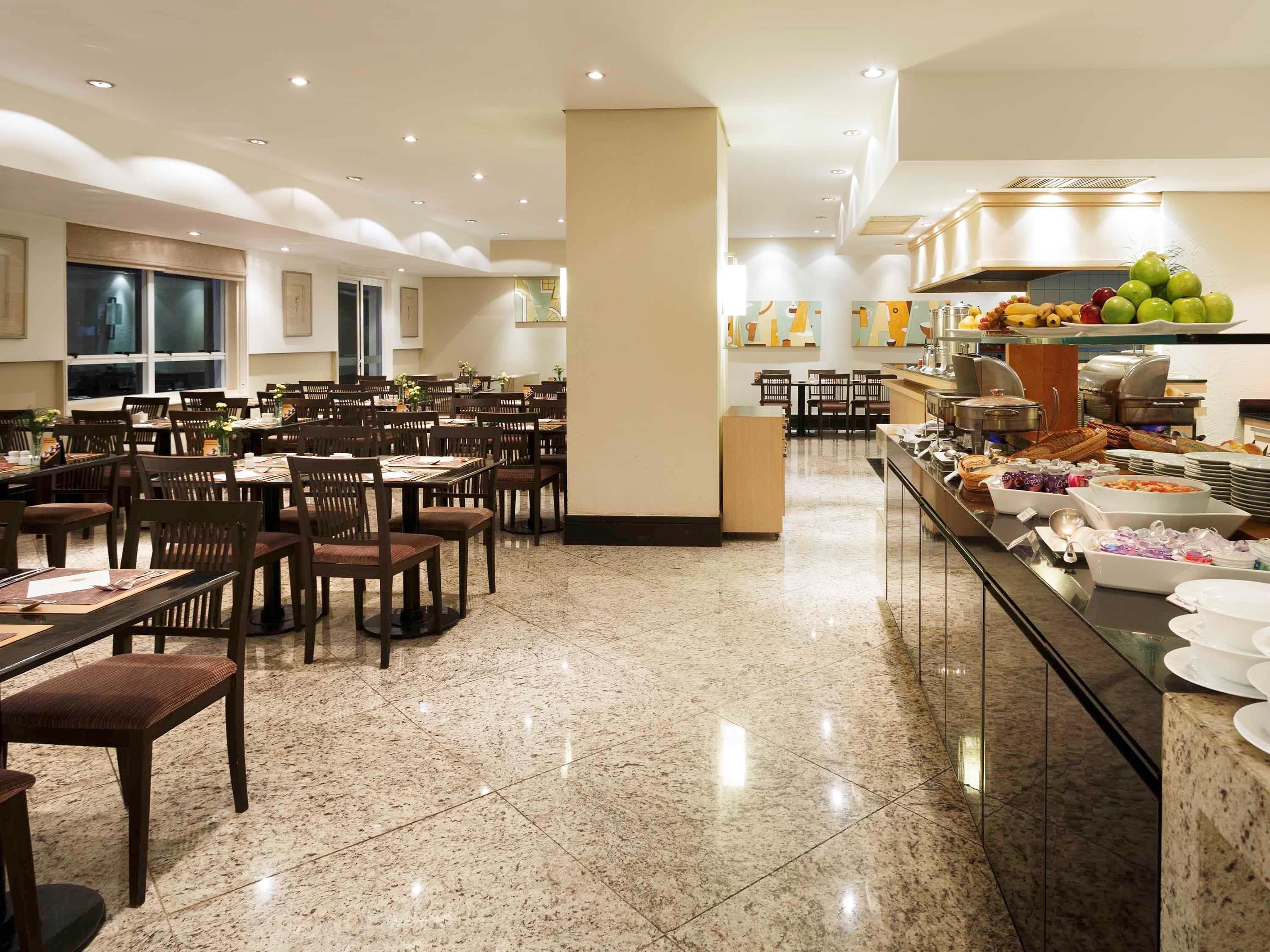 Uma sala de jantar ampla com várias mesas e cadeiras, um buffet com diversos alimentos e um grande pilar no centro.