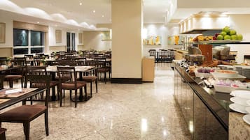 Uma sala de jantar ampla com várias mesas e cadeiras, um buffet com diversos alimentos e um grande pilar no centro.