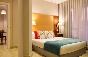 Quarto de hotel com cama, abajures, mesa de cabeceira e vista da cidade através de cortinas leves.