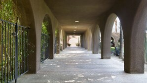 Uma galeria coberta com arcos, piso de concreto e uma cerca de metal de um lado.