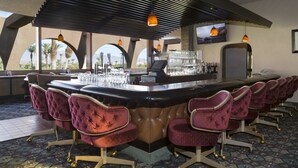 Uma área de bar com balcão, banquetas e vista para palmeiras através de janelas em arco.