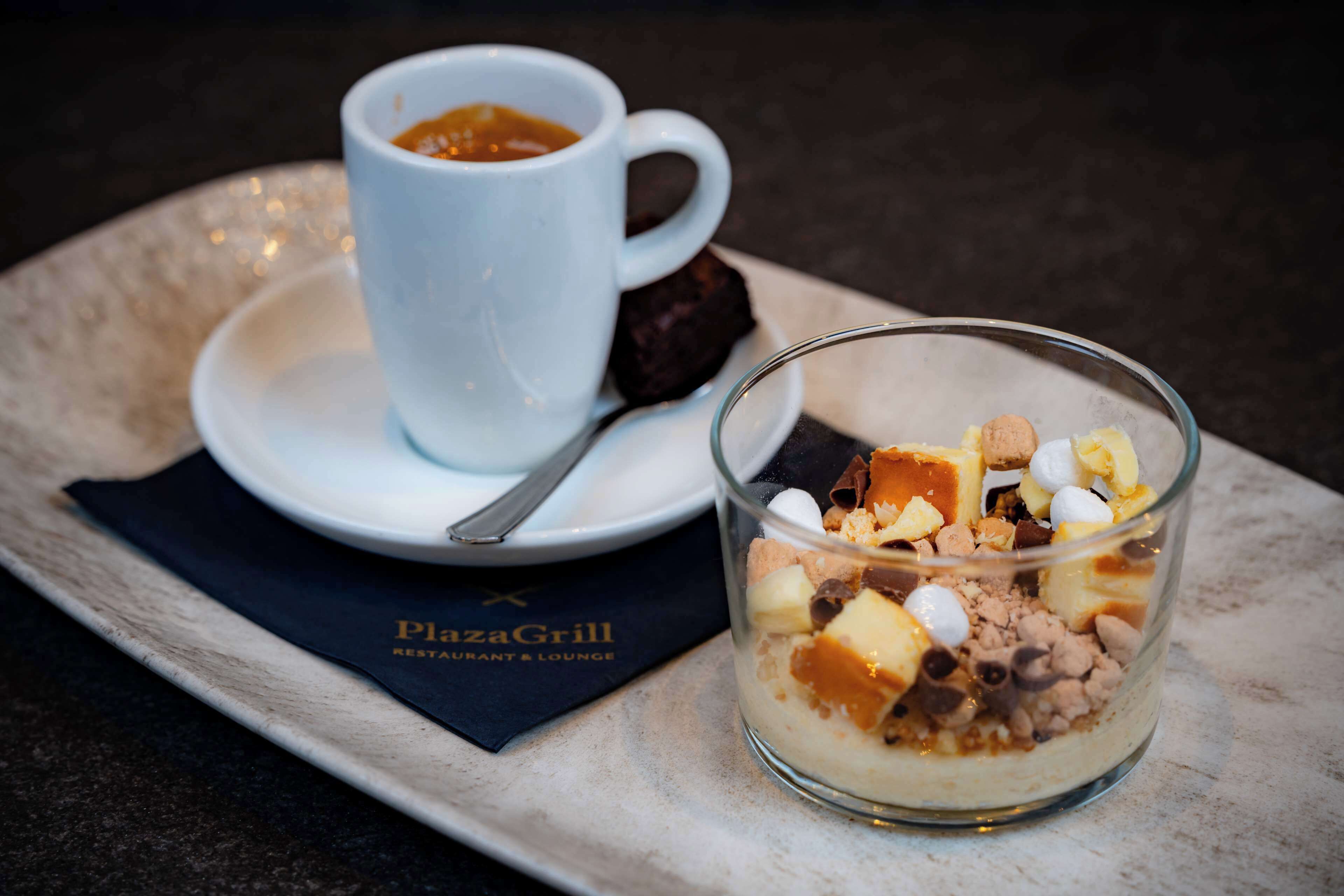 Um copo com sobremesa em camadas, uma chávena de café e um brownie de chocolate sobre um tabuleiro.