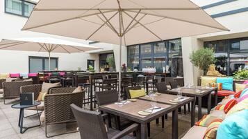 Uma área de refeições ao ar livre com móveis de vime, grandes guarda-sóis e vista para o restaurante.
