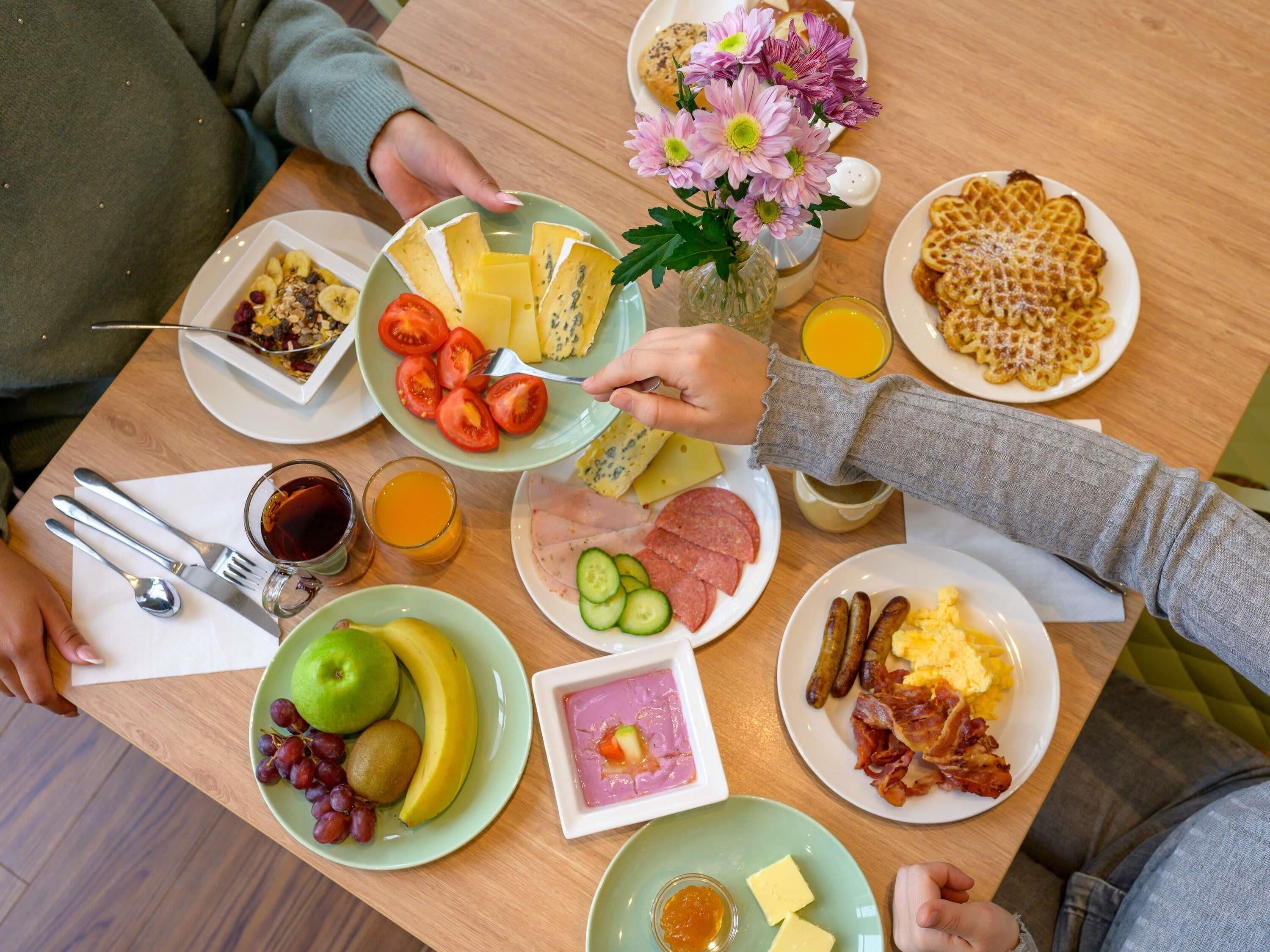 Uma mesa de café da manhã com diversos pratos, incluindo ovos, salsichas, presunto, queijo e frutas.