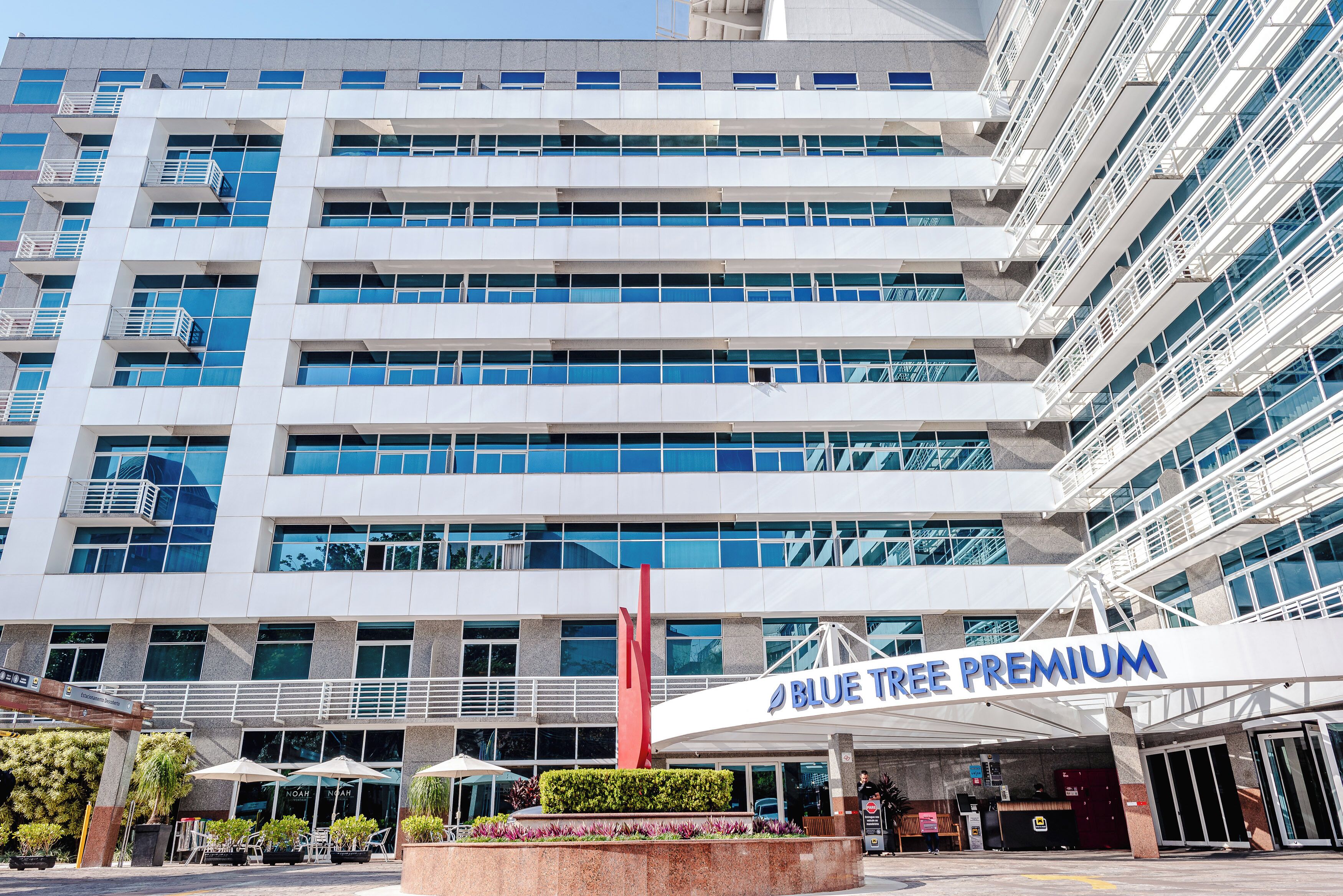 Um hotel de vários andares com fachada moderna, entrada destacada com a placa "BLUE TREE PREMIUM" e área externa com mesas e guarda-sóis.
