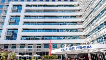 Um hotel de vários andares com fachada moderna, entrada destacada com a placa "BLUE TREE PREMIUM" e área externa com mesas e guarda-sóis.