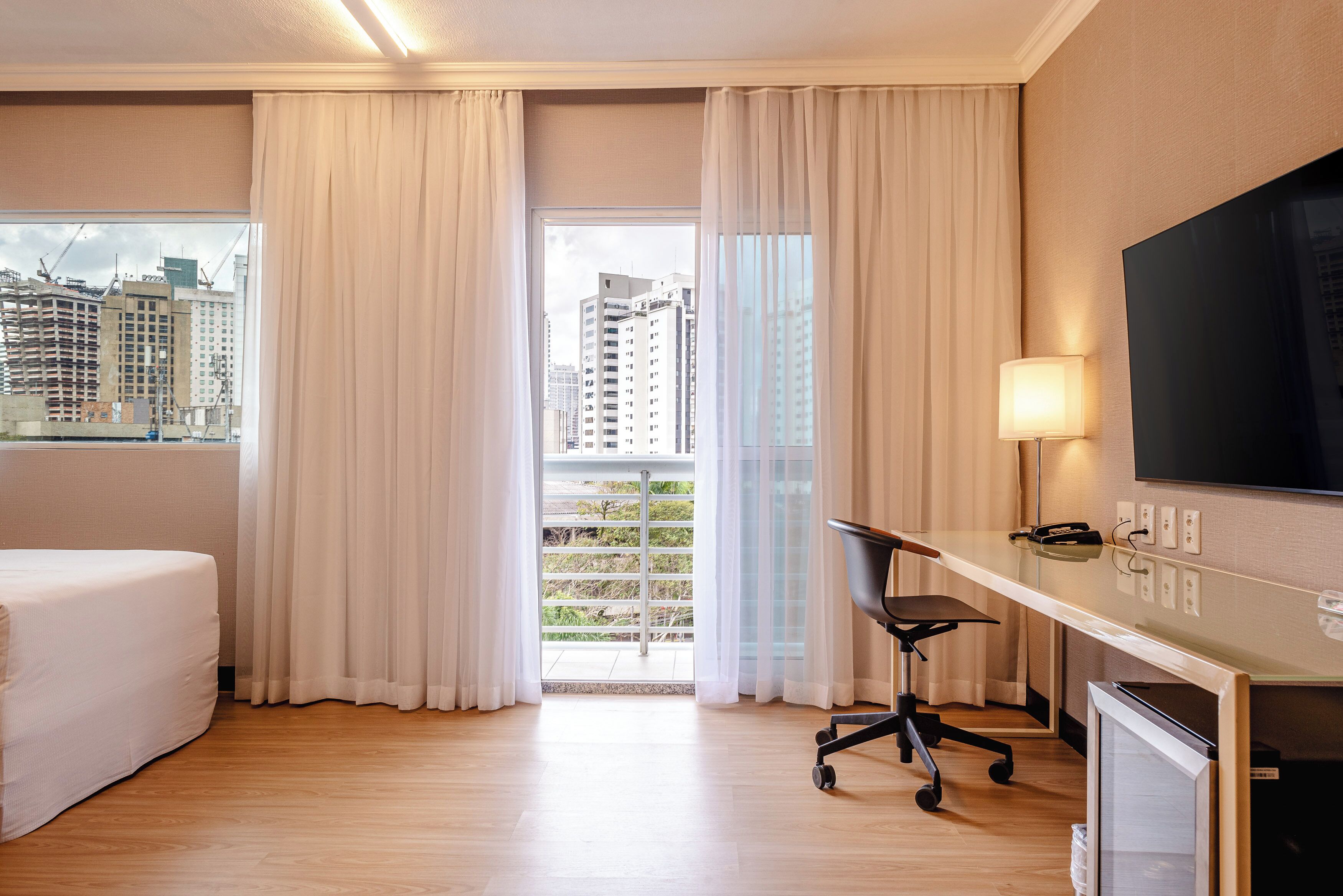 Quarto de hotel com uma cama, uma escrivaninha com cadeira, televisão e vista para edifícios pela janela.