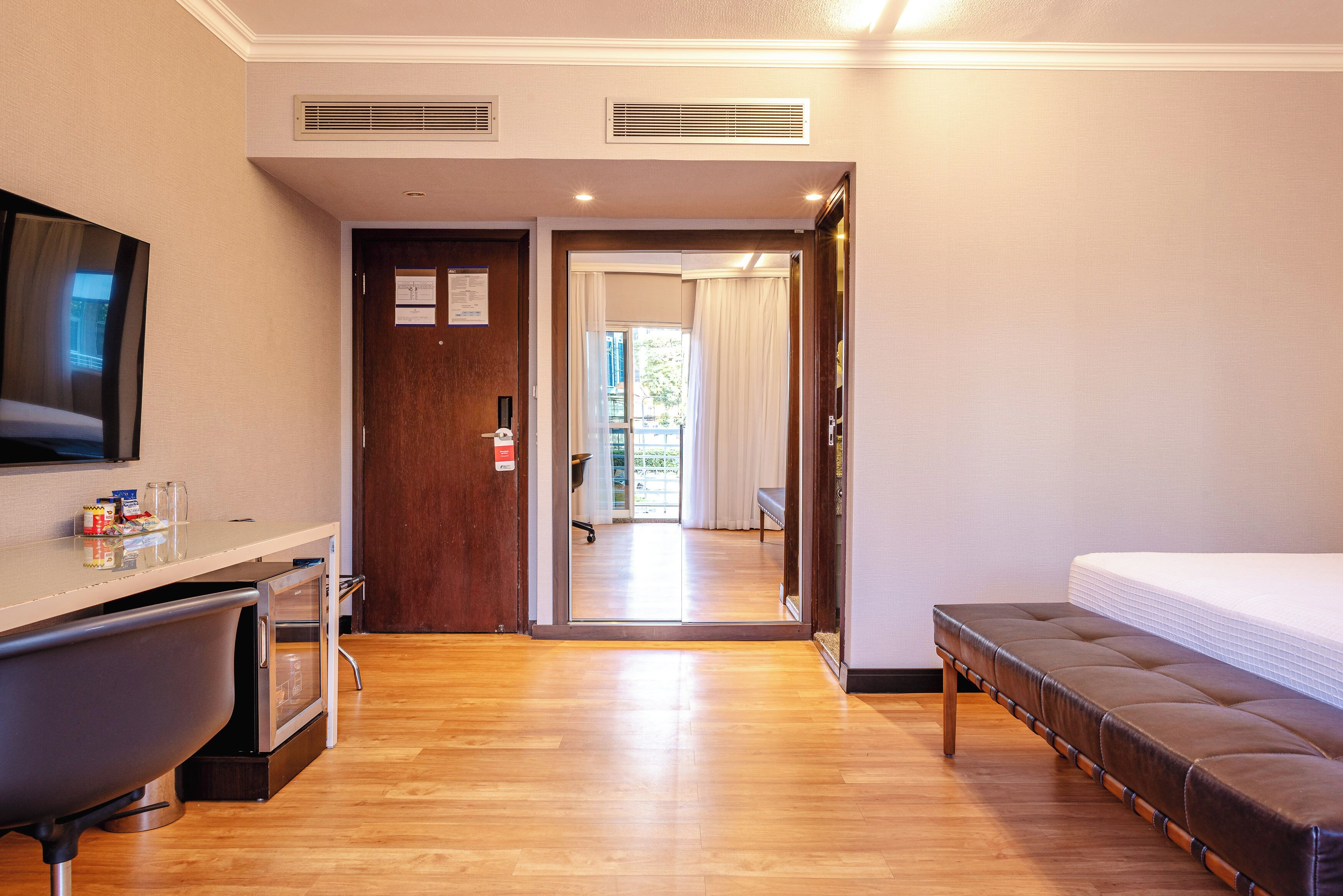 Quarto de hotel com piso de madeira, um sofá, uma TV, uma mesa de escritório com computador e máquina de café, e uma porta que dá para uma varanda.
