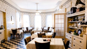Uma sala de jantar com piso xadrez, várias mesas e cadeiras, e um armário decorativo com diversos objetos.