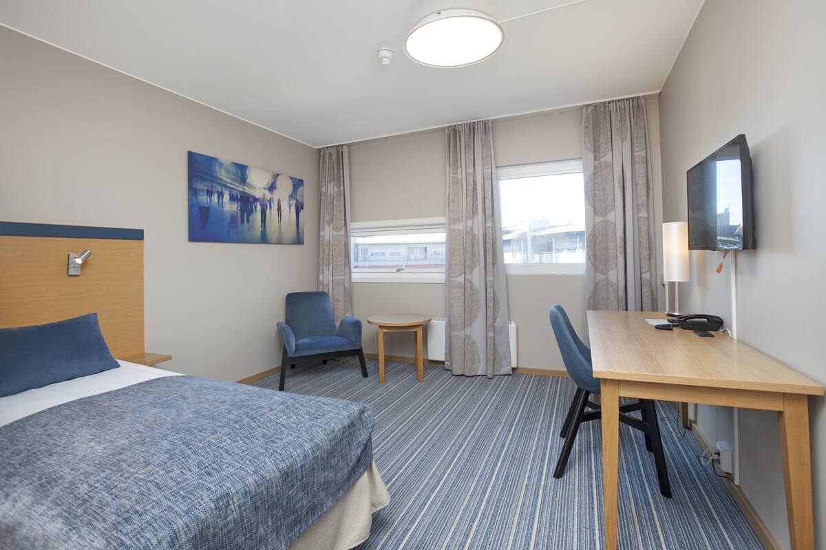 Quarto de hotel com cama, escrivaninha, cadeira, televisão e janela com cortinas.