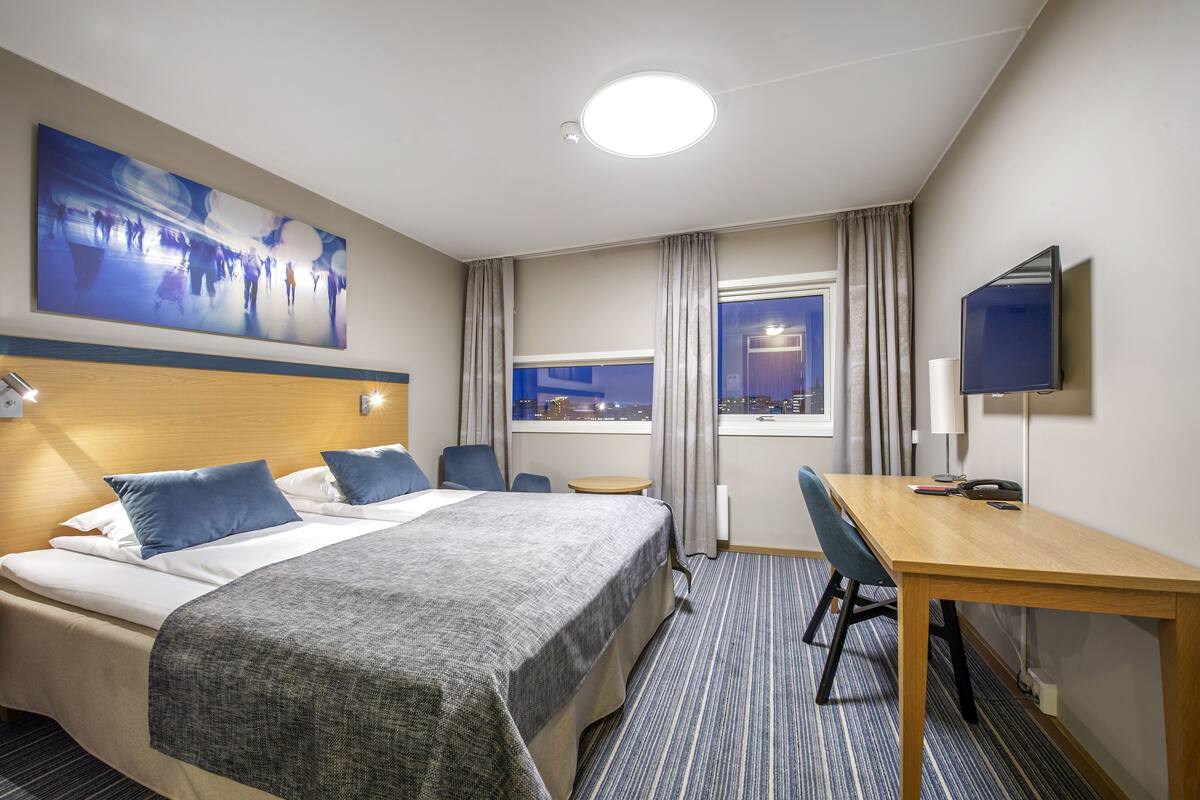 Quarto de hotel com uma cama grande, uma escrivaninha, uma cadeira, uma televisão e vista para a cidade pela janela.