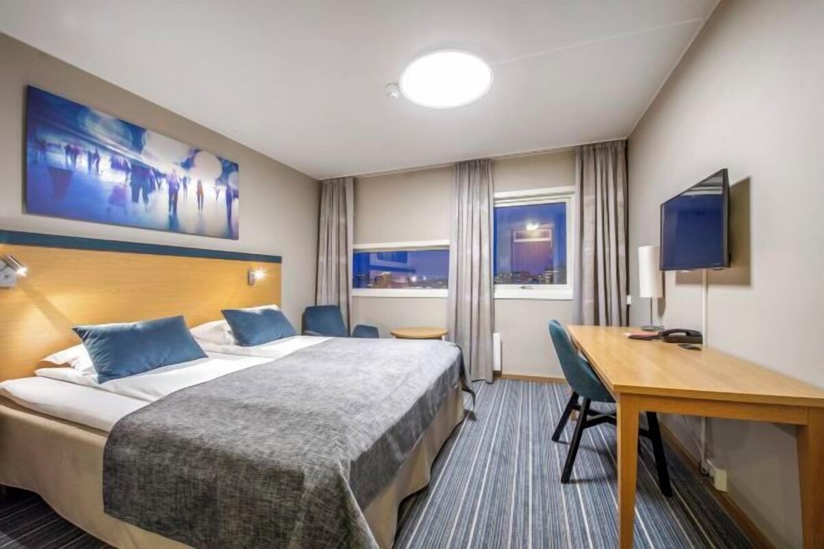 Quarto de hotel com cama, escrivaninha, cadeira, TV e janela com cortinas.