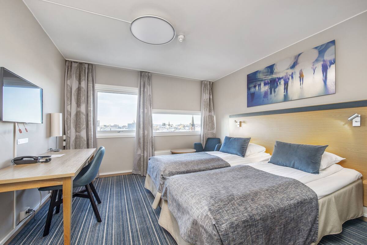 Quarto de hotel com duas camas, uma escrivaninha, uma cadeira e uma janela ampla com vista para a cidade.