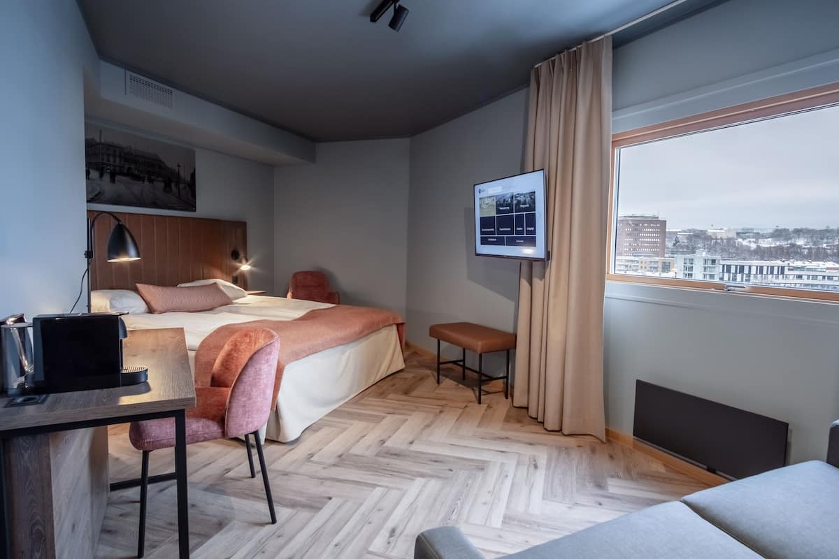 Quarto de hotel moderno com cama, escrivaninha, cadeira e uma grande janela com vista para a cidade.