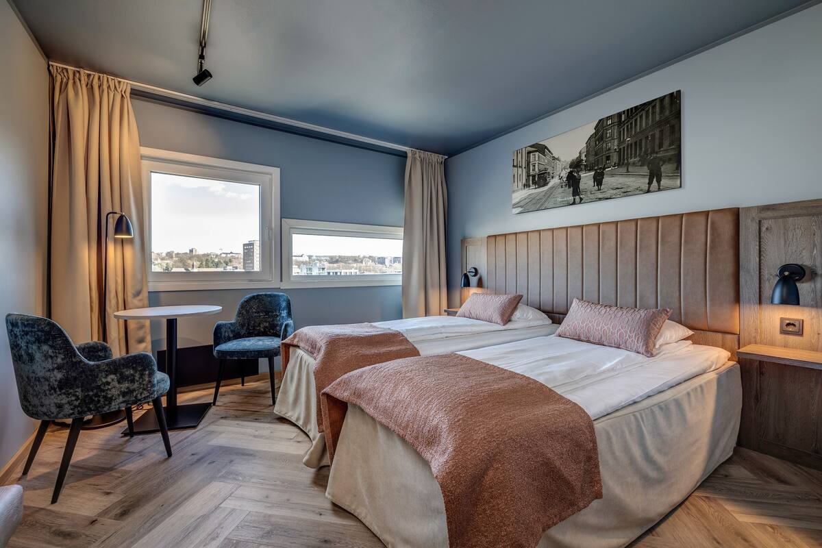 Quarto de hotel com duas camas, uma mesa redonda, duas cadeiras, uma janela com vista e um quadro pendurado na parede.
