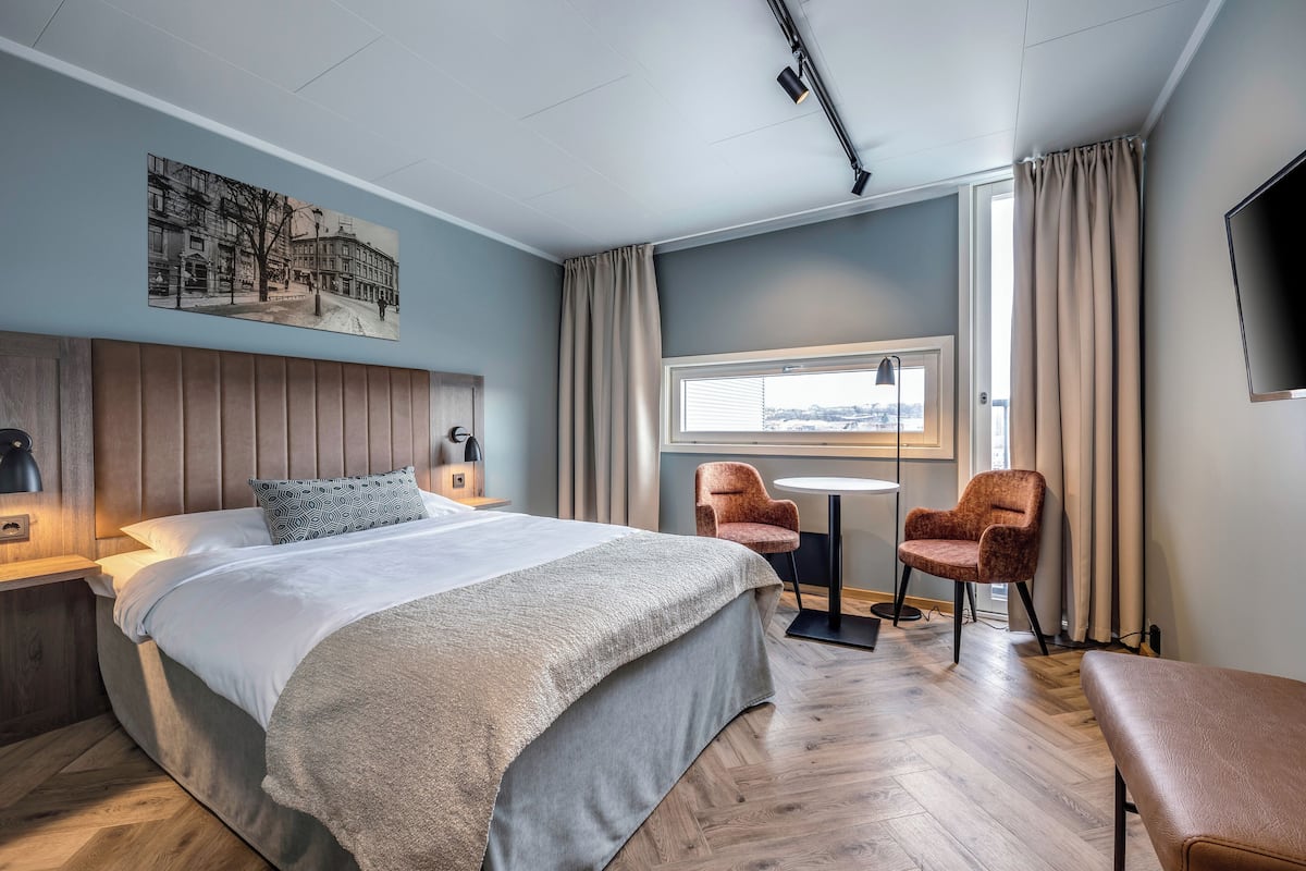Quarto de hotel com uma cama grande, cabeceira de madeira, uma mesa pequena com duas cadeiras, um banco, uma janela com cortinas e uma televisão de tela plana fixada na parede.