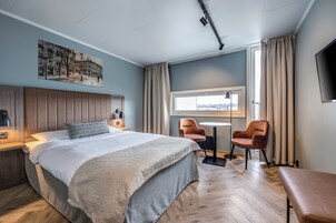 Quarto de hotel com uma cama grande, cabeceira de madeira, uma mesa pequena com duas cadeiras, um banco, uma janela com cortinas e uma televisão de tela plana fixada na parede.