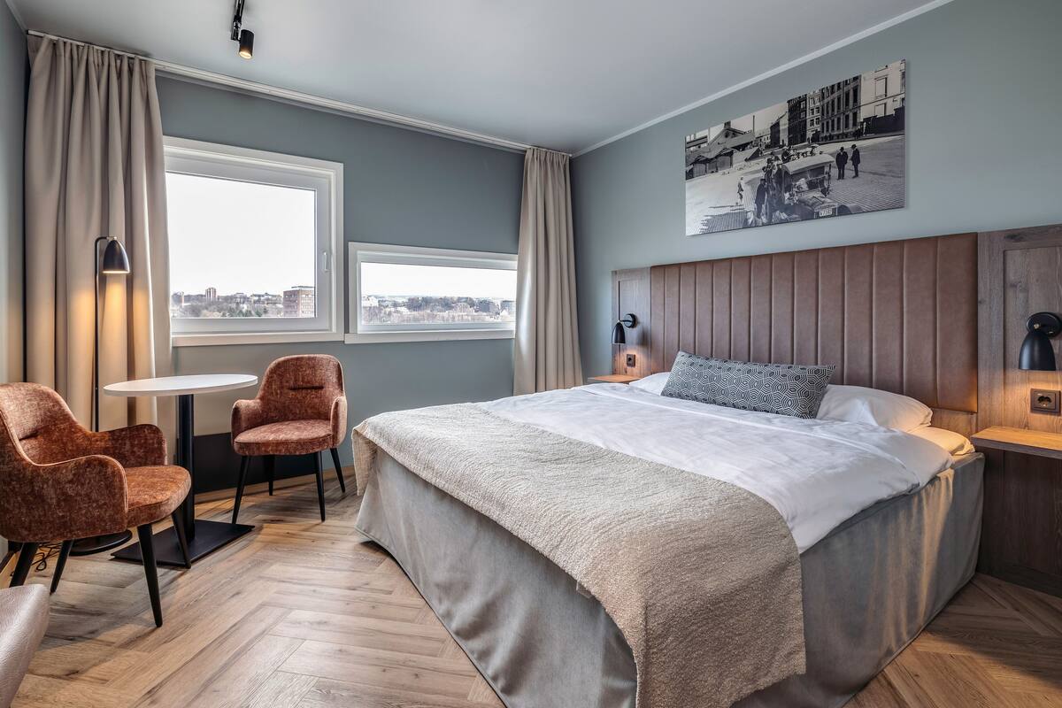 Quarto de hotel com uma cama grande, duas poltronas, uma mesinha e vista para a cidade pela janela.