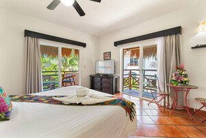 Quarto de hotel com cama, televisão, varanda com mesa e cadeiras, e vista para o verde lá fora.