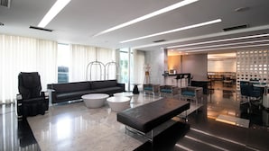 Um lobby de hotel moderno com um sofá preto, cadeiras brancas e azuis, um banco e uma área de refeições com parede estampada.