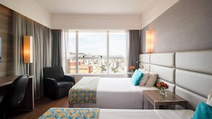 Quarto de hotel com uma cama grande, uma escrivaninha, uma cadeira, uma mesa pequena e vista para a cidade através de grandes janelas.