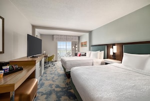 Quarto de hotel com duas camas, uma mesa, uma televisão e uma janela com vista.