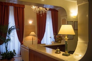 Uma área de recepção de hotel com balcão de mármore, luminária dourada, cortina vermelha e um candelabro.