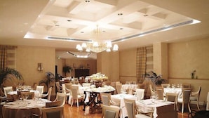 Uma sala de jantar ampla com várias mesas redondas preparadas para refeições, um lustre central e plantas em vasos.