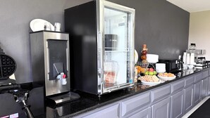 Uma área de cozinha com geladeira, grill para waffles, cafeteira e uma fruteira.