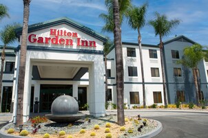 Exterior do Hilton Garden Inn, com grande cobertura de entrada, palmeiras e um elemento decorativo de água.