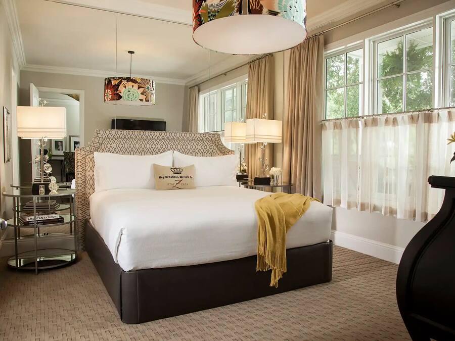 Quarto de hotel com uma cama grande, duas luminárias de cabeceira, um espelho, cômoda e uma janela com cortinas.
