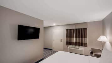 Quarto de hotel com cama, escrivaninha e cadeira, televisão na parede, porta e janela com cortinas.