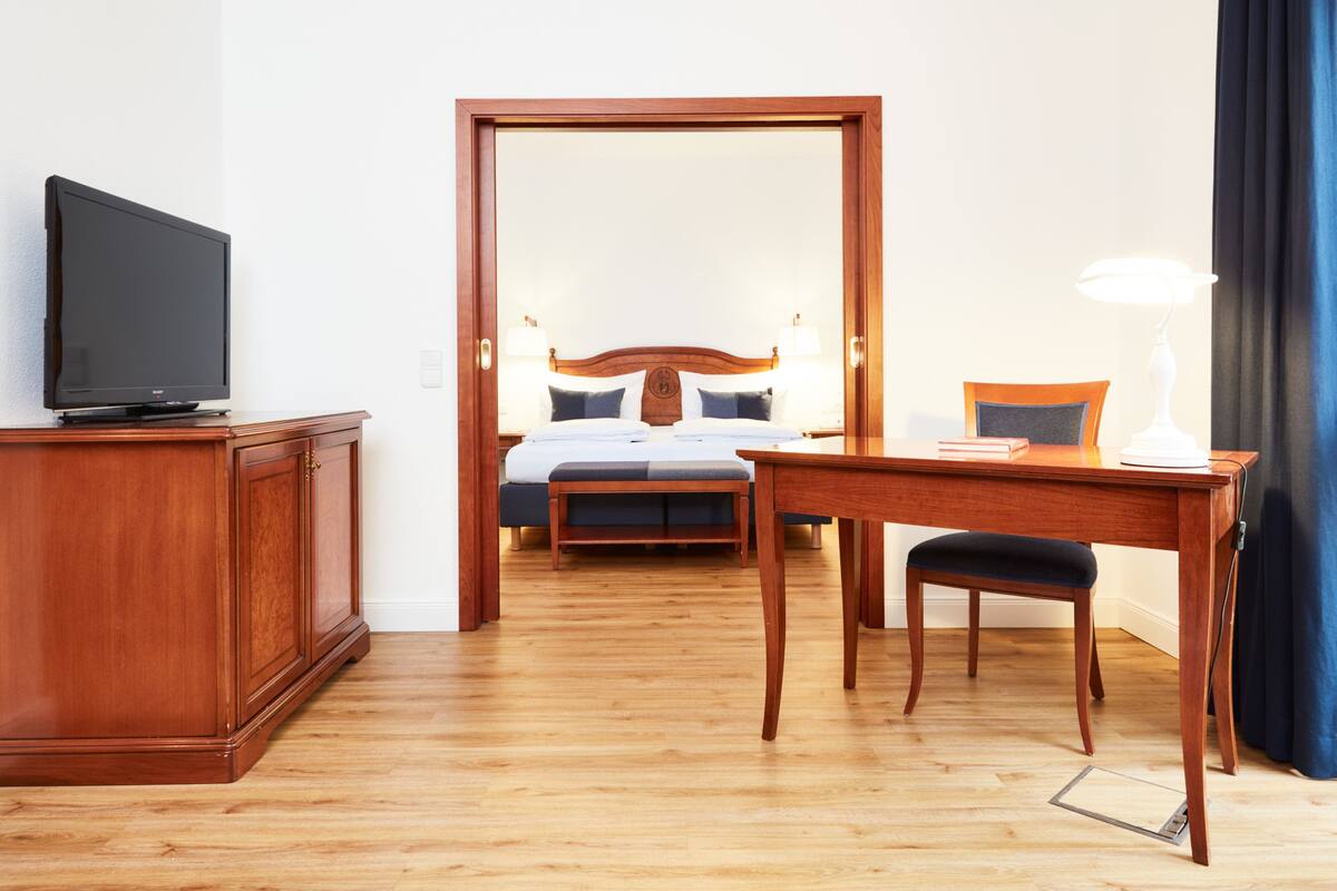Quarto de hotel com escrivaninha de madeira, televisão, cama com cabeceira e mesa de cabeceira.