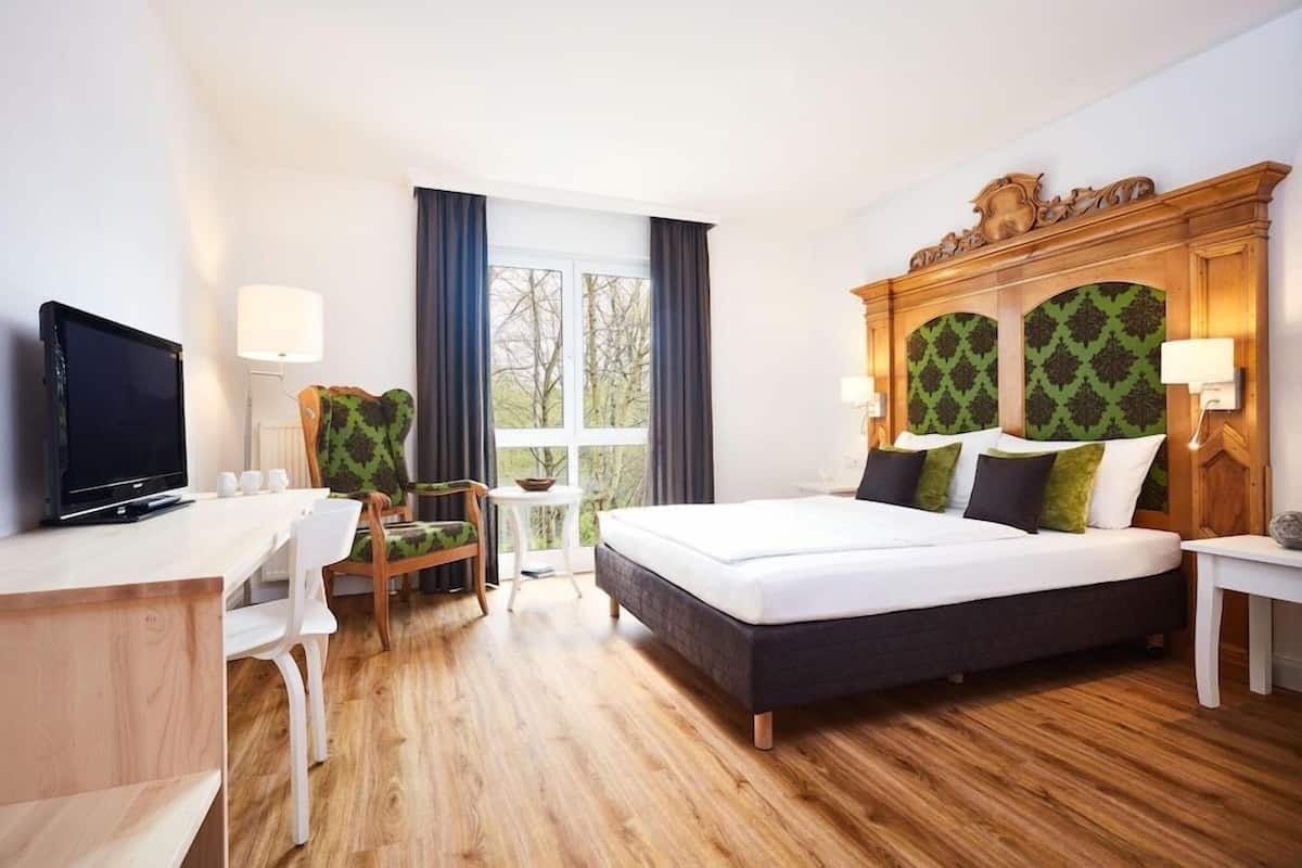Quarto de hotel com cama, escrivaninha, cadeira, televisão e janela com cortinas.