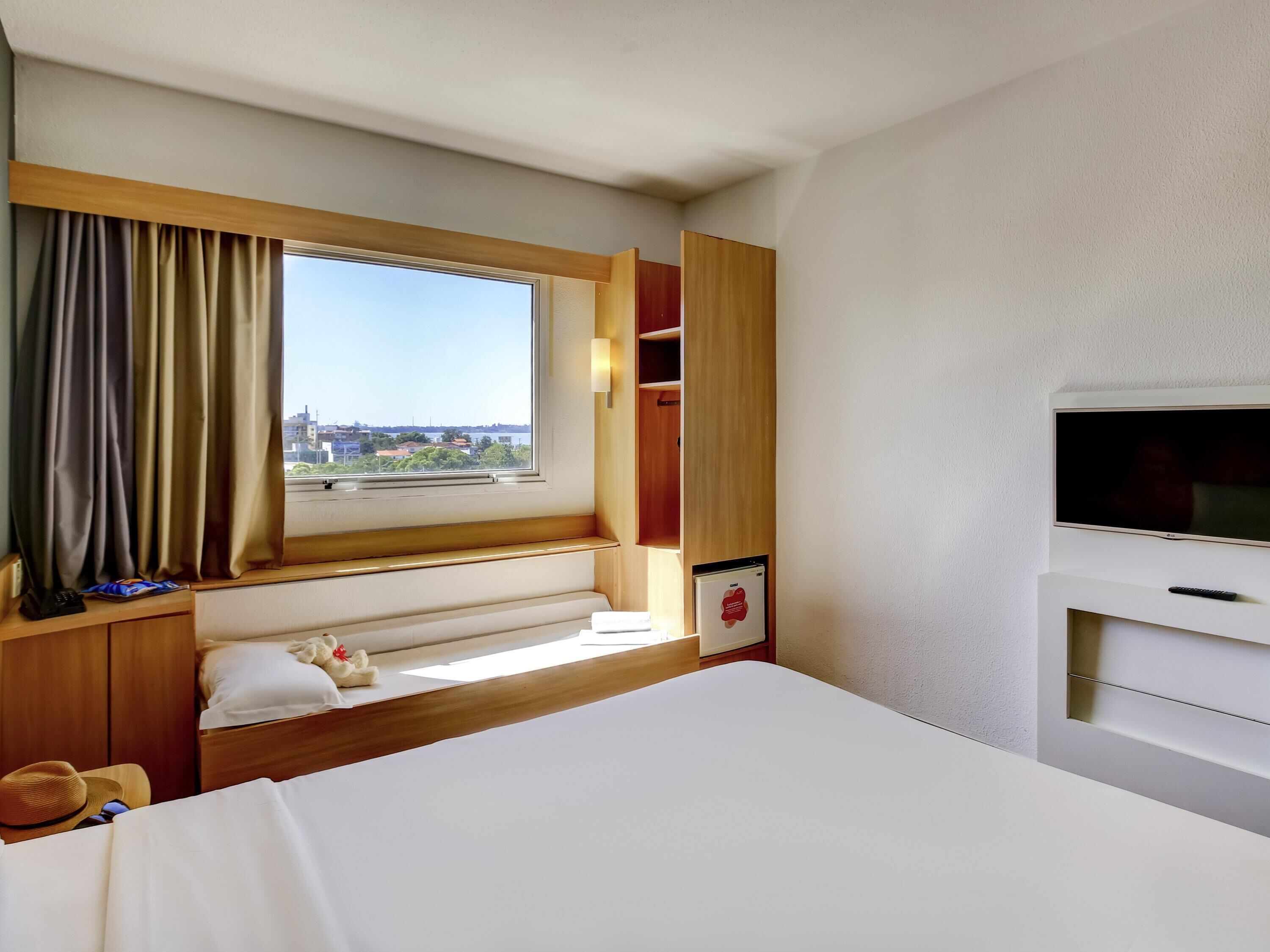 Quarto de hotel com cama, televisão, janela com cortinas e vista da cidade.
