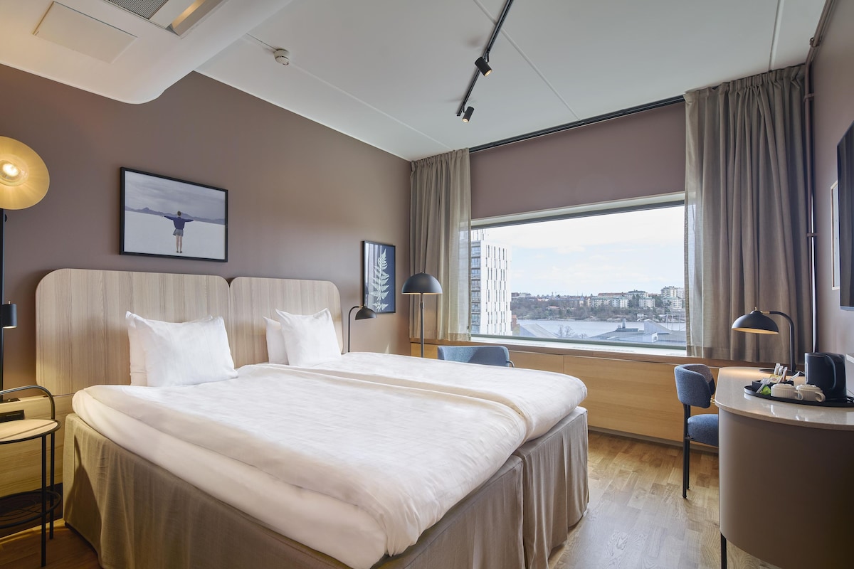 Quarto de hotel com uma cama grande, uma escrivaninha, uma cadeira, uma luminária e uma janela com vista para a cidade.