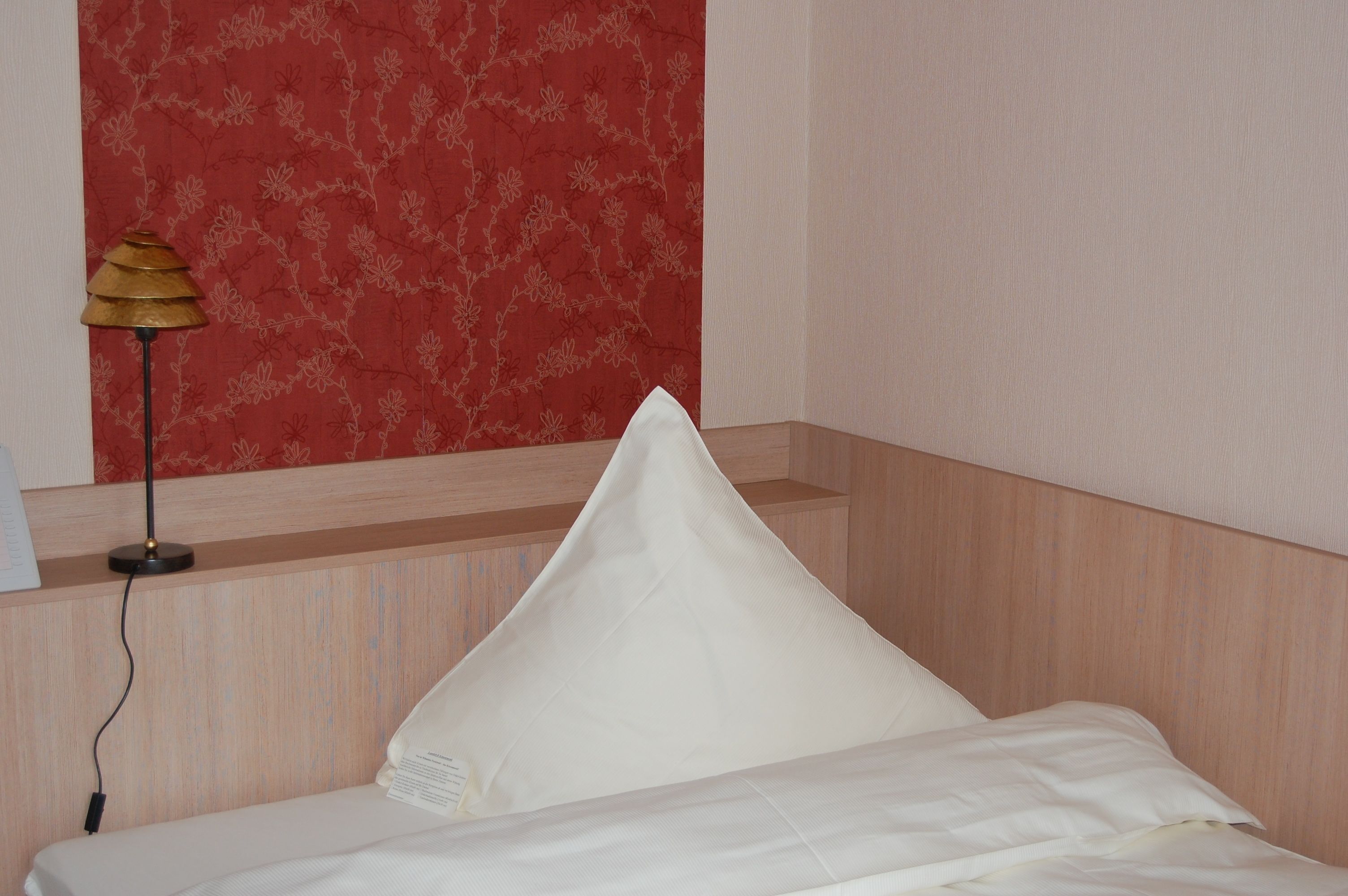 Cama bem arrumada com travesseiro branco e abajur sobre um criado-mudo de madeira.