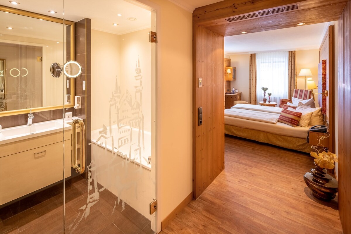 Quarto de hotel com piso de madeira, cama com travesseiros, mesa de cabeceira com abajur e banheiro com espelho e toucador.